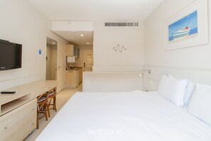 Quarto de hotel com uma cama, uma televisão, uma escrivaninha com cadeiras e uma cozinha compacta ao fundo.