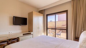 Quarto de hotel com cama, televisão, guarda-roupa, secretária e varanda com cadeiras.