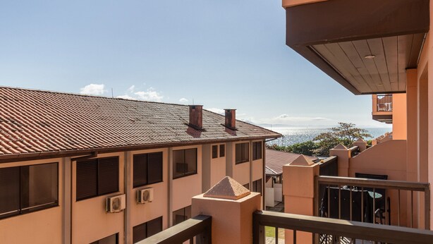 Um edifício residencial com telhado de telhas, várias janelas e uma varanda com vista para a paisagem urbana e montanhas ao longe.