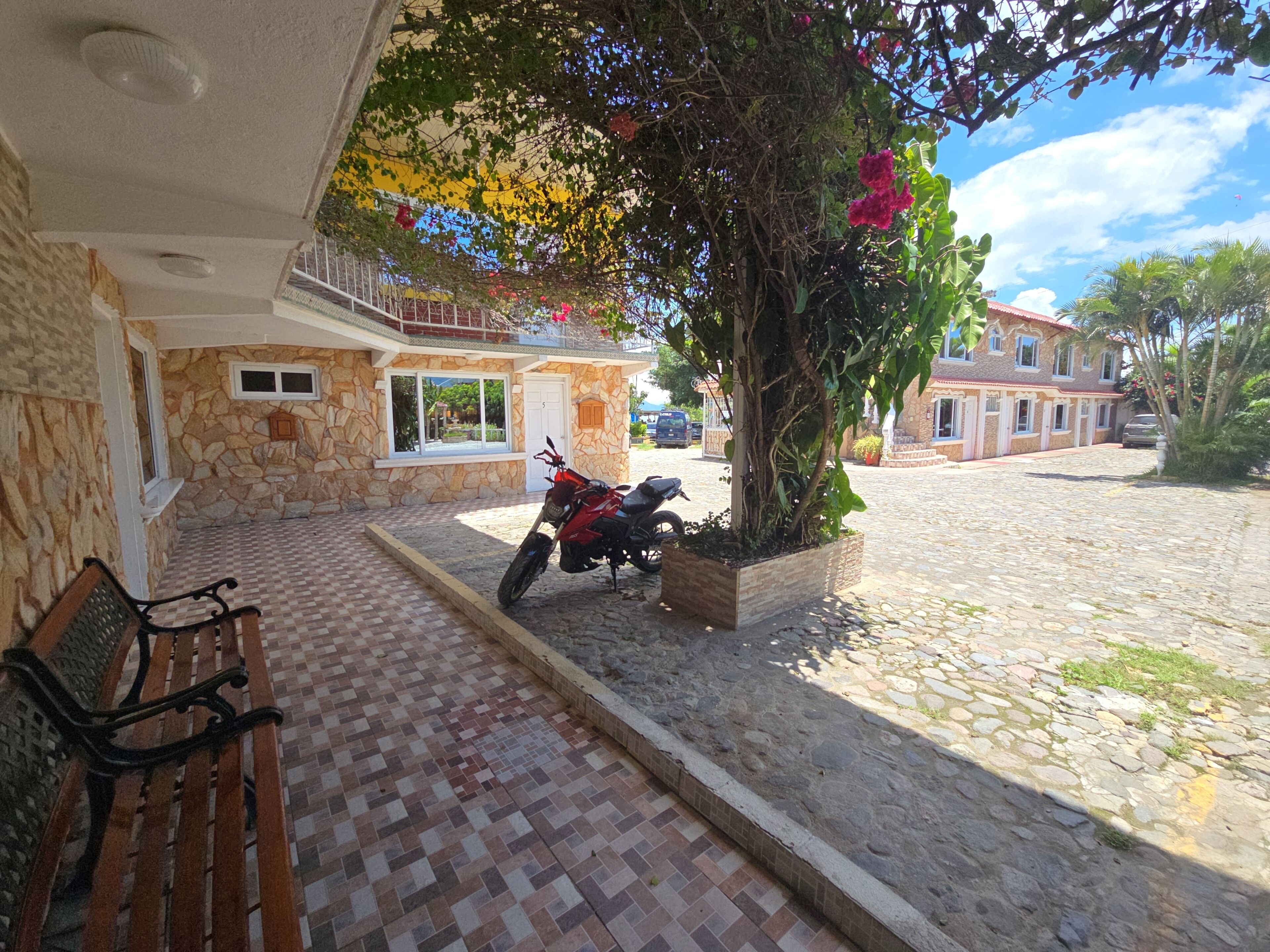 Um pátio pavimentado com um banco, uma motocicleta e uma árvore com flores cor-de-rosa.