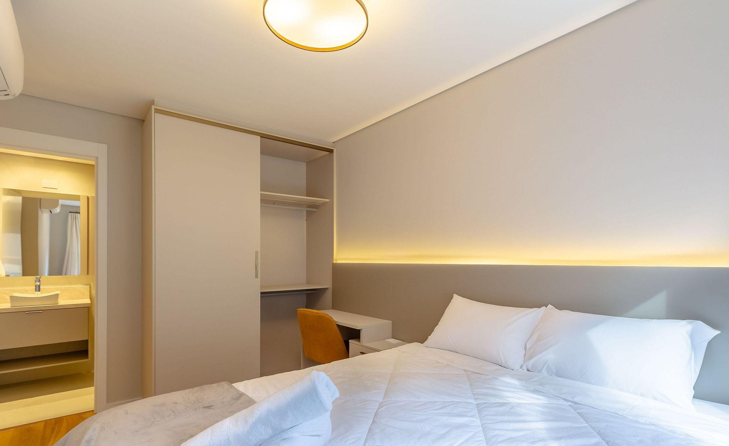 Quarto de hotel com cama, escrivaninha, cadeira, guarda-roupa e espelho.