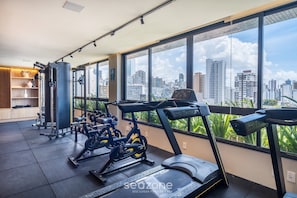 Uma academia com equipamentos de ginástica, grandes janelas e vista para a cidade.