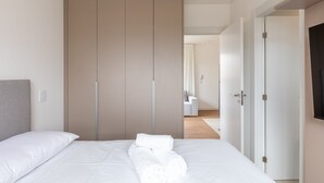 Cama bem arrumada com roupa de cama e toalhas brancas, cabeceira cinza e um guarda-roupa grande com portas deslizantes.