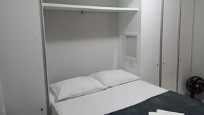 Uma cama de solteiro com roupa de cama branca, um cobreleito de cor escura e uma pequena mesa de cabeceira com um ventilador.