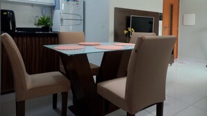 Uma sala de jantar com mesa de vidro, quatro cadeiras, geladeira, cafeteira e uma TV ao fundo.