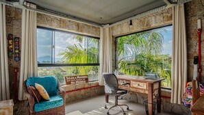 Quarto com parede de pedra, cadeira de vime com almofadas, escrivaninha com cadeira e amplas janelas com vista para o verde.