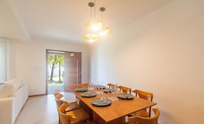 Uma sala de jantar com uma mesa de madeira posta para seis pessoas, rodeada por seis cadeiras de madeira. Há uma porta que dá para o exterior, e um sofá branco é visível ao fundo.