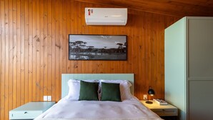 Quarto de hotel com parede revestida em madeira, cama com roupa de cama branca e verde, ar condicionado fixado, quadro emoldurado, mesa de cabeceira com abajur e criado-mudo com um livro.