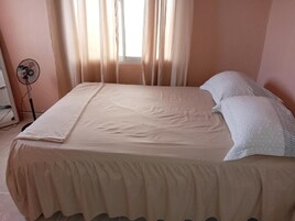 Uma cama de solteiro com colcha e almofadas bege, um ventilador e uma janela com cortinas leves.