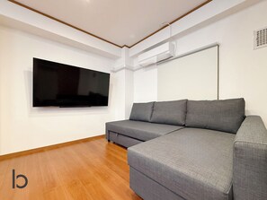 Uma sala de estar moderna com um sofá cinza, uma televisão de tela plana fixada na parede e piso de madeira.