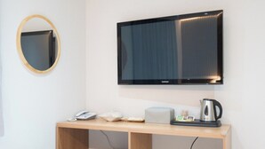 A hotel room with a flat-screen TV mounted on the wall, a wooden desk with a phone, a kettle, and a small speaker.