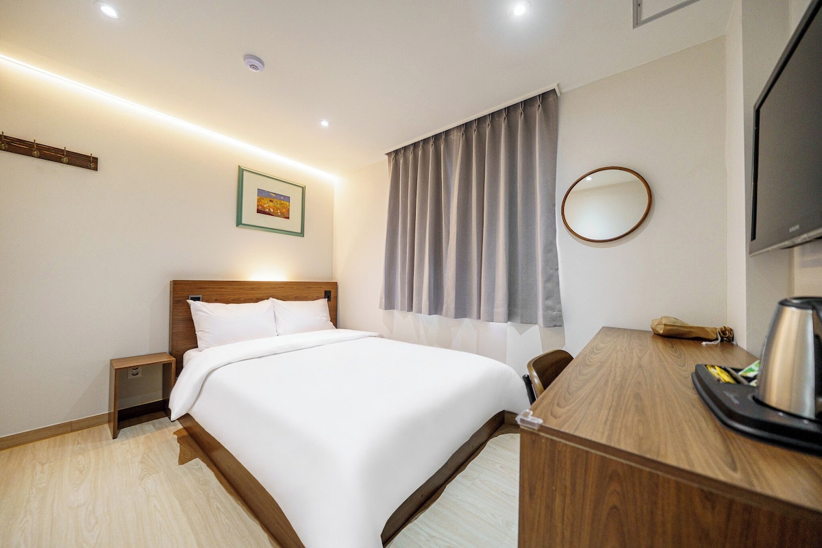 Quarto de hotel com cama, mesa de cabeceira, escrivaninha com chaleira, televisão, janela com cortinas e um quadro na parede.