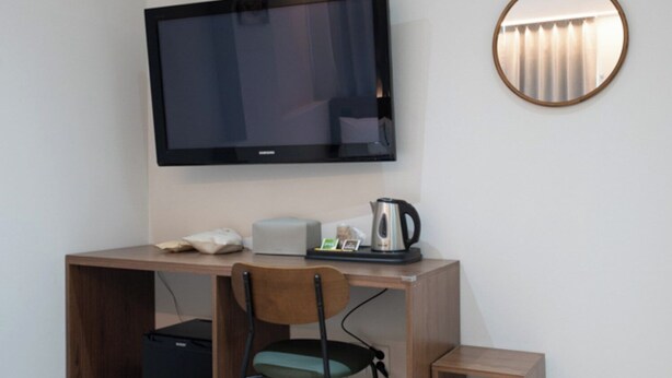A hotel room with a flat-screen TV mounted on the wall, a desk with a chair, a small cabinet, a kettle, and a bedside table.