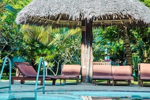 Área de piscina com espreguiçadeiras e uma cobertura de palha.