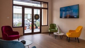 Um saguão de hotel com uma grande porta de vidro, uma televisão presa na parede e três cadeiras.