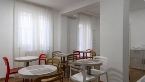 Uma área de refeições com várias mesas e cadeiras, uma cozinha com armários e uma janela com cortinas.