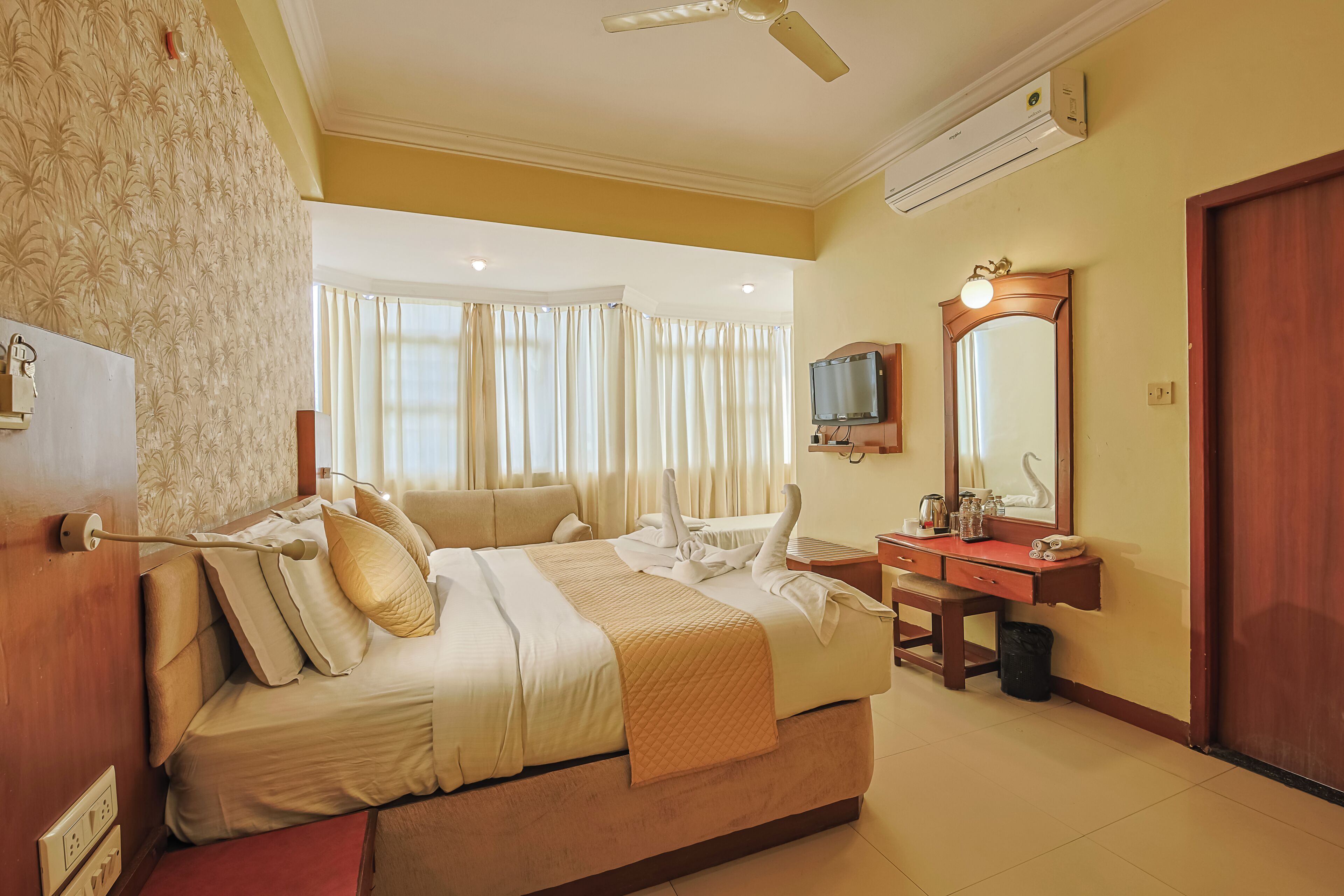 Quarto de hotel com cama, escrivaninha, espelho, televisão e ar condicionado.