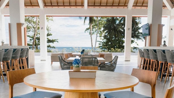 Uma área de refeições ao ar livre espaçosa, com uma mesa redonda rodeada por cadeiras de madeira com almofadas azuis, com vista para uma paisagem deslumbrante do oceano e palmeiras.