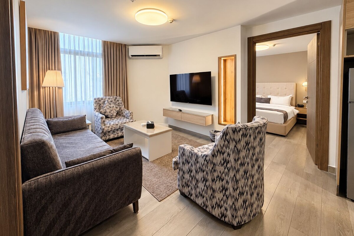 Quarto de hotel moderno com sofá, poltrona, televisão, mesa de cabeceira e cama com encosto.
