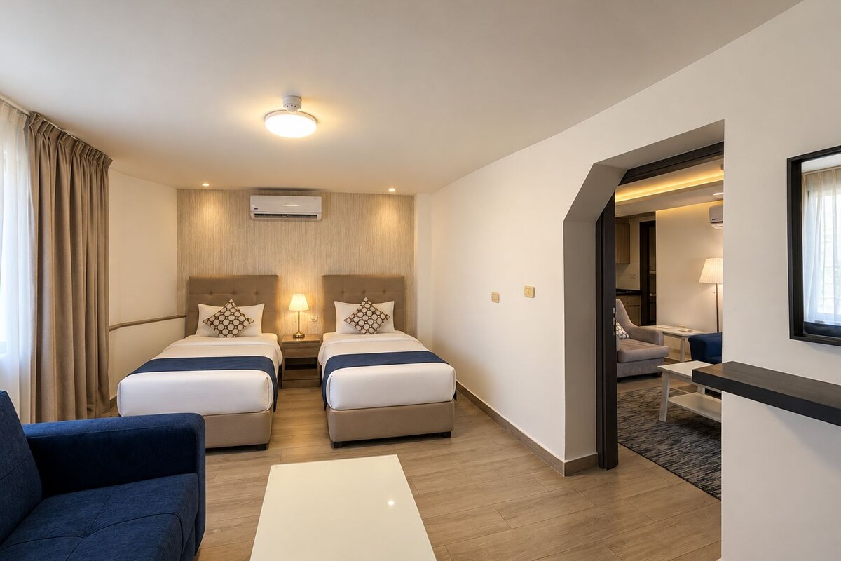 Quarto de hotel com duas camas, um sofá, uma escrivaninha e um banheiro visível por uma porta aberta.