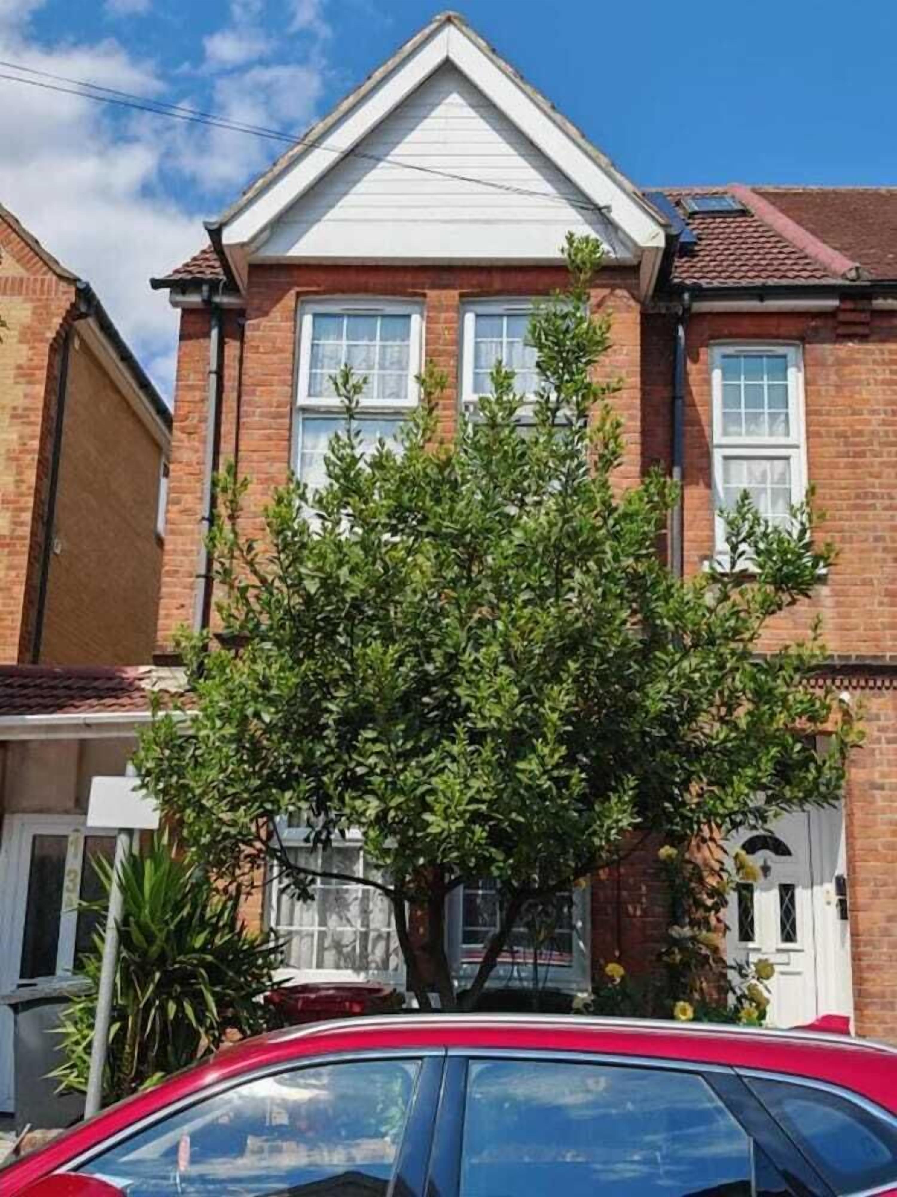 Uma casa de dois andares de tijolo com porta e janelas brancas, uma árvore na frente e um carro vermelho estacionado.