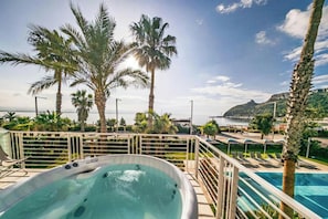 Uma banheira de hidromassagem rodeada por palmeiras, com vista para uma piscina e uma paisagem deslumbrante de um corpo d'água e colinas ao longe.
