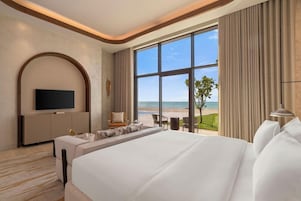 Quarto de hotel com uma cama grande, televisão, área de estar e vista para a praia.