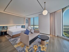 Quarto de hotel moderno com uma cama grande, um sofá, uma mesinha e vista para o aeroporto.
