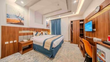 Quarto de hotel com cama, mesa de cabeceira, televisão e escrivaninha com cadeira.