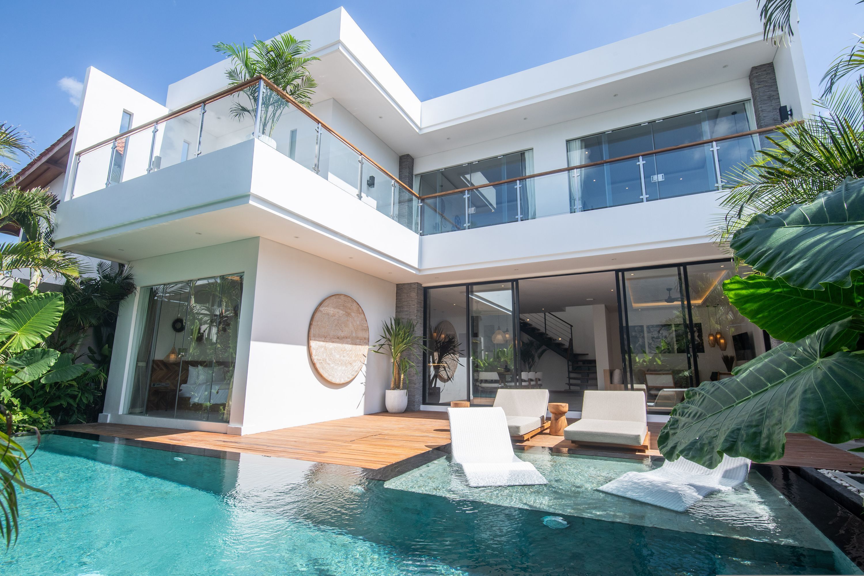 Uma casa moderna de dois andares com varanda de vidro, piscina e plantas tropicais.