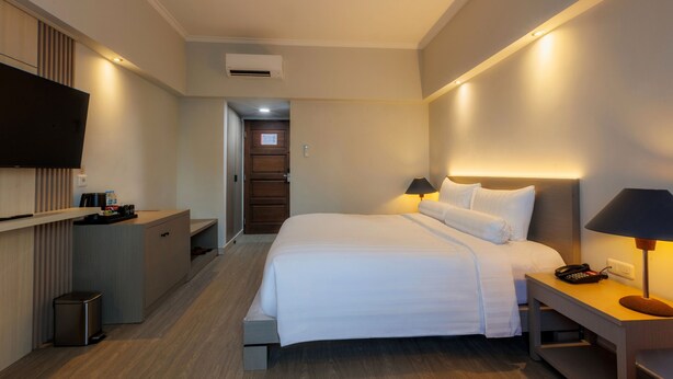 Quarto de hotel com cama, mesas de cabeceira, televisão e escrivaninha com computador.