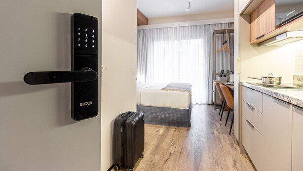 Quarto de hotel moderno com fechadura digital, uma cama, pequeno espaço de cozinha e uma mala.
