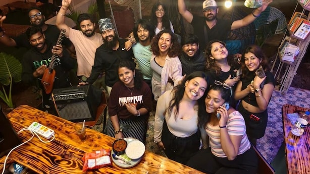 Um grupo de pessoas reunido em um bar, com uma mesa de madeira, algumas segurando bebidas e um aparelho de som visível ao fundo.