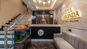 Um hall de hotel com escadaria, balcão de recepção, estátua de Buda, relógio e placa com a inscrição 'HOTEL CHARONDA'.