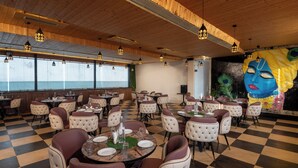 Restaurante com piso xadrez, mesas redondas com cadeiras, iluminação pendente e um mural na parede.