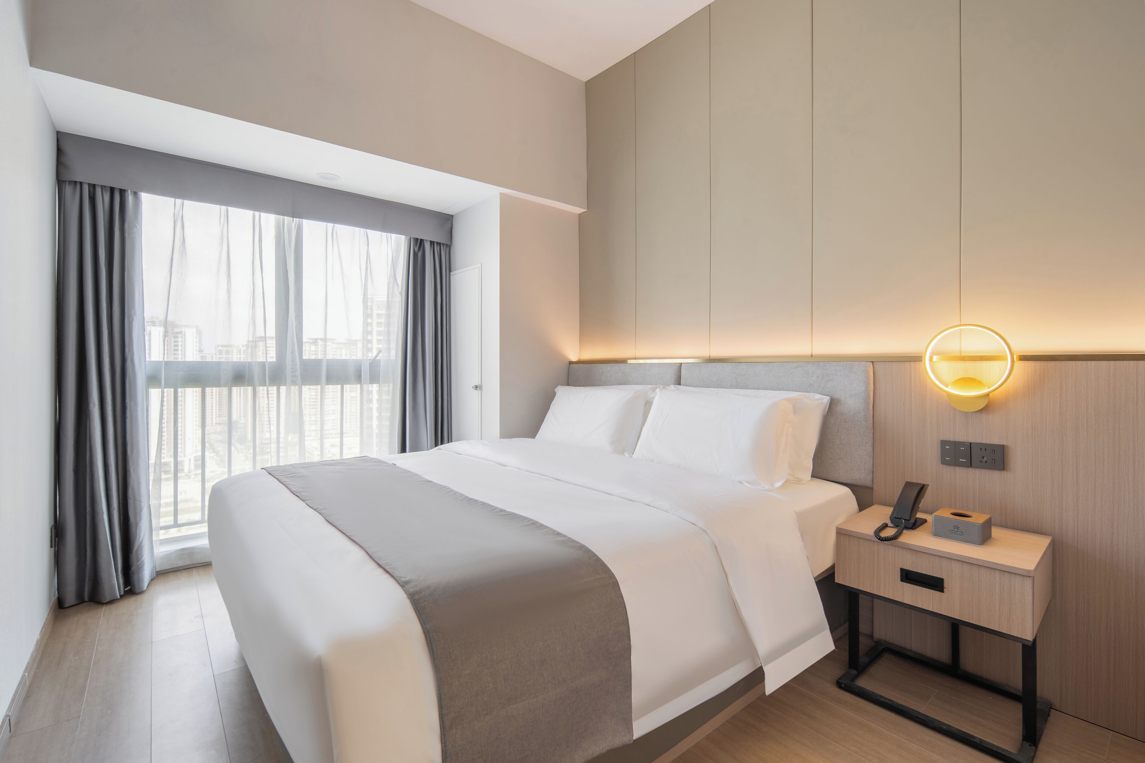 Quarto de hotel com uma cama grande, uma mesa de cabeceira com abajur e vista para edifícios pela janela.