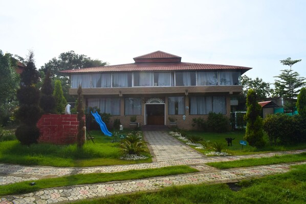 Uma casa de dois andares com telhado de telhas vermelhas, entrada frontal e um pequeno jardim com um escorrega azul.