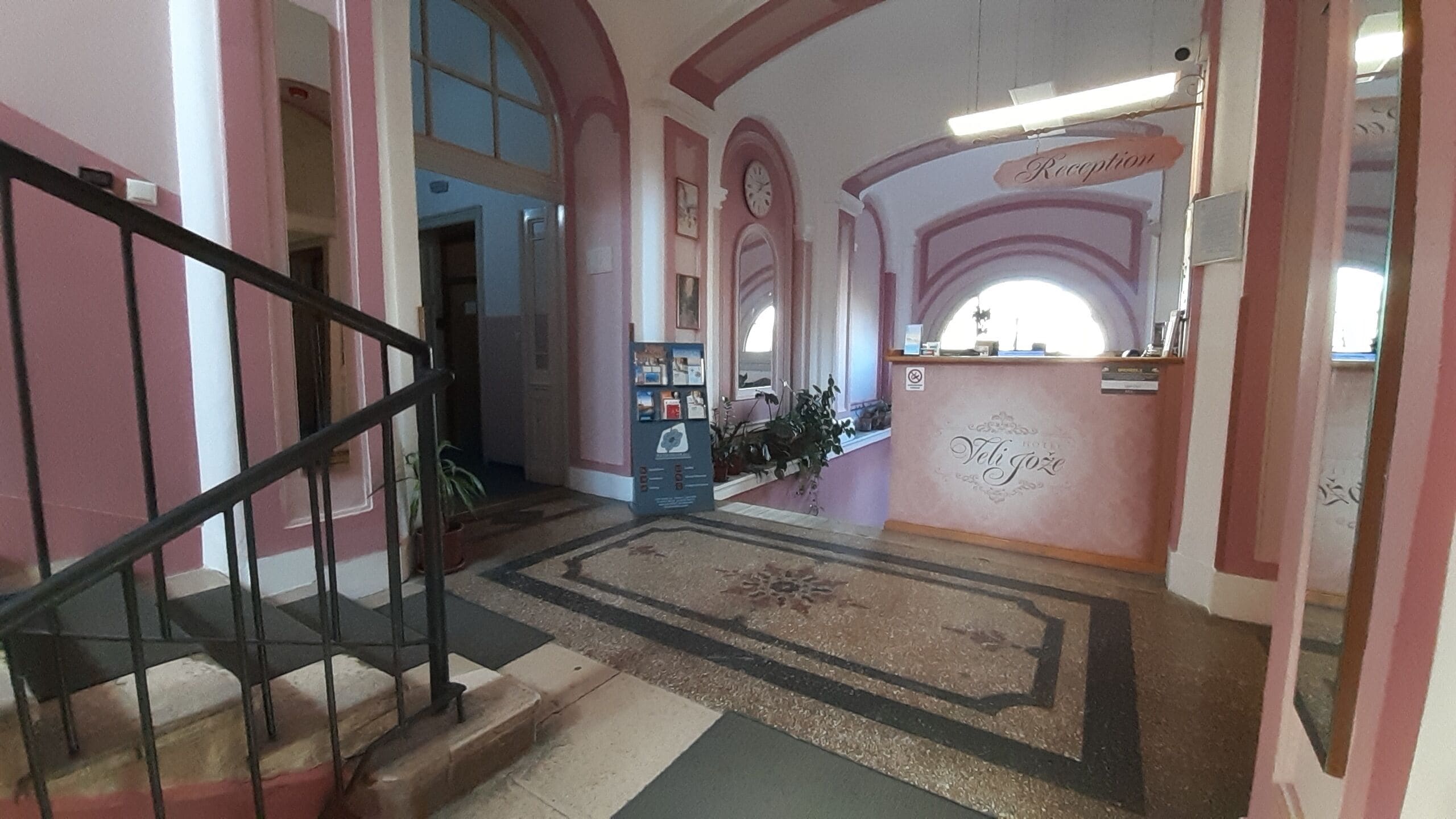 A hotel reception area with a reception desk, a staircase, and a wall with a clock.