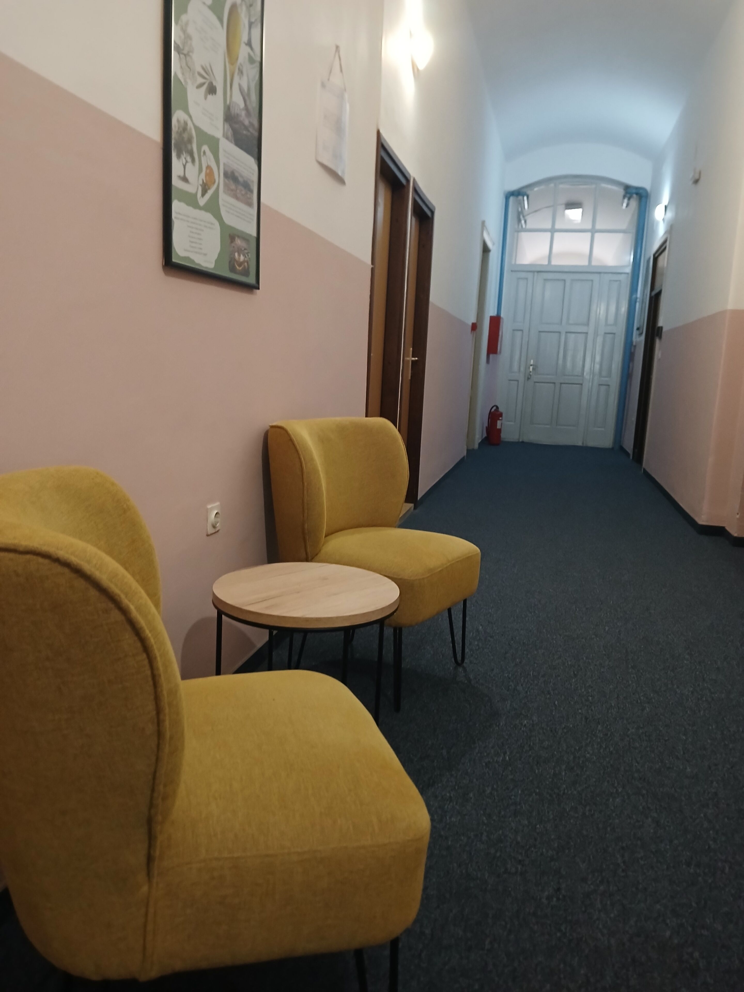 Um corredor com duas poltronas amarelas e uma pequena mesa redonda, um cartaz informativo emoldurado na parede, e uma porta no final.