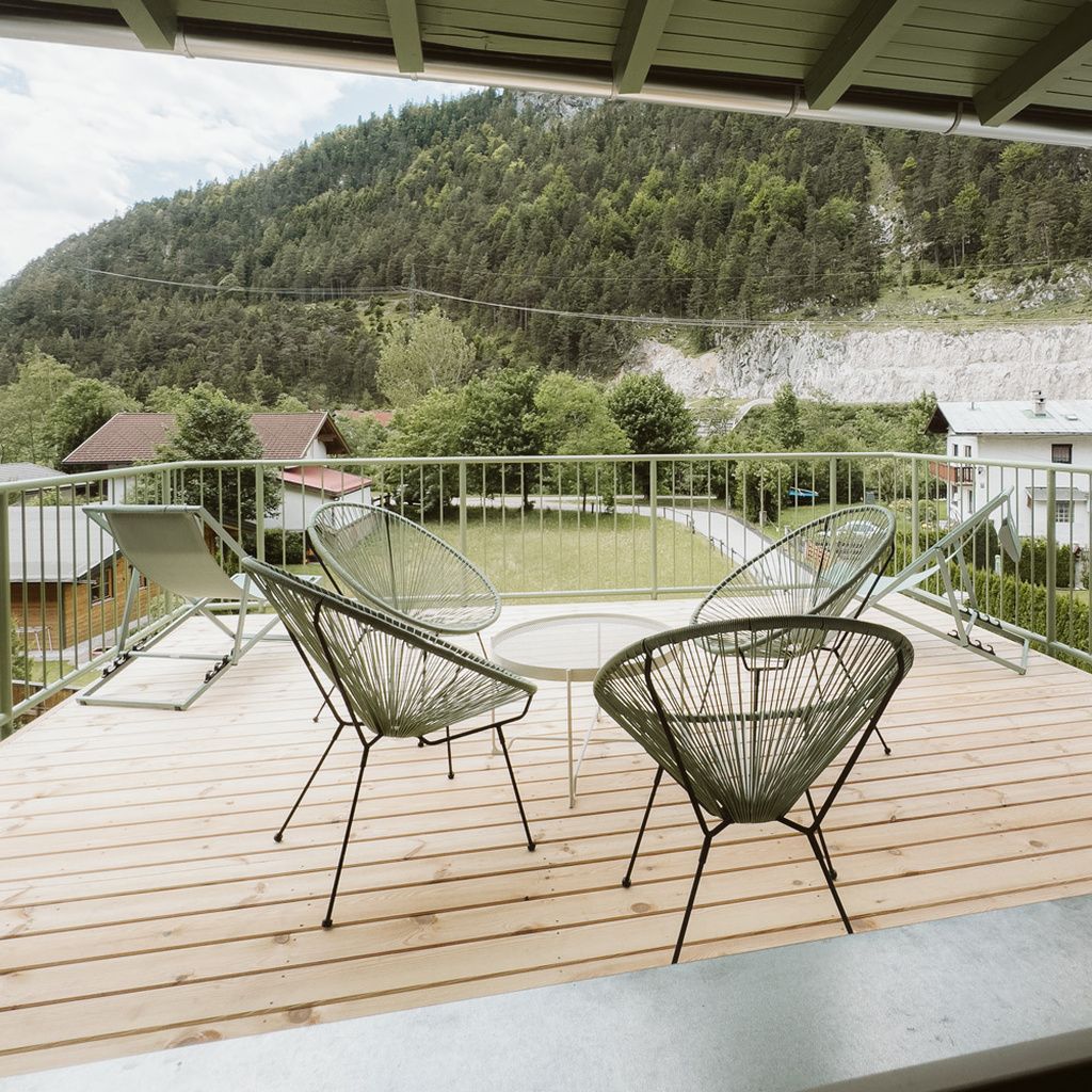 Um deck de madeira com duas cadeiras de metal e uma mesa, com vista para uma encosta arborizada.