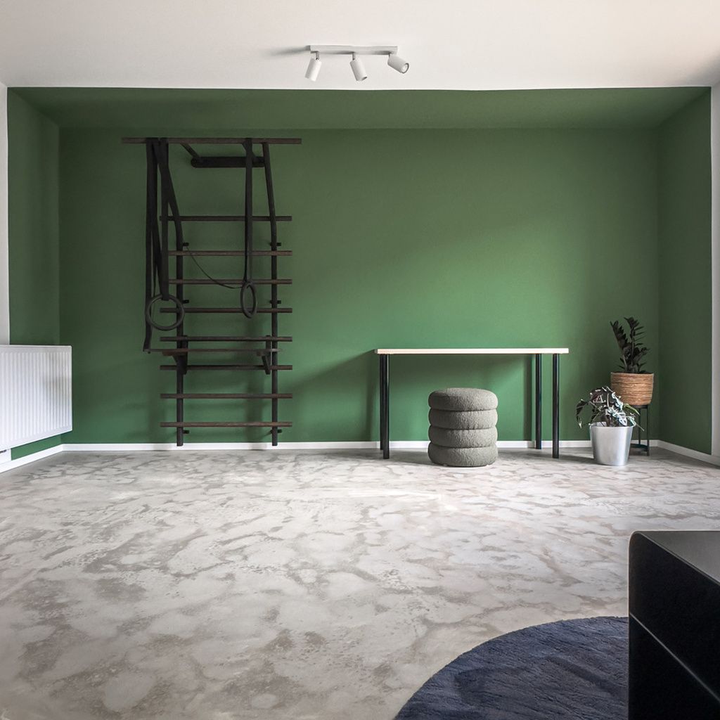 Quarto com parede verde, luminária branca no teto, escada de metal preta, mesa branca, otomano cinza e planta em vaso.