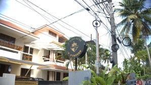 Placa de hotel com emblema circular, um edifício de vários andares com varandas e um ambiente tropical com palmeiras.