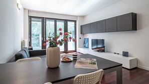 Uma sala de estar moderna com área de jantar, grandes janelas, televisão de tela plana e um vaso com flores.