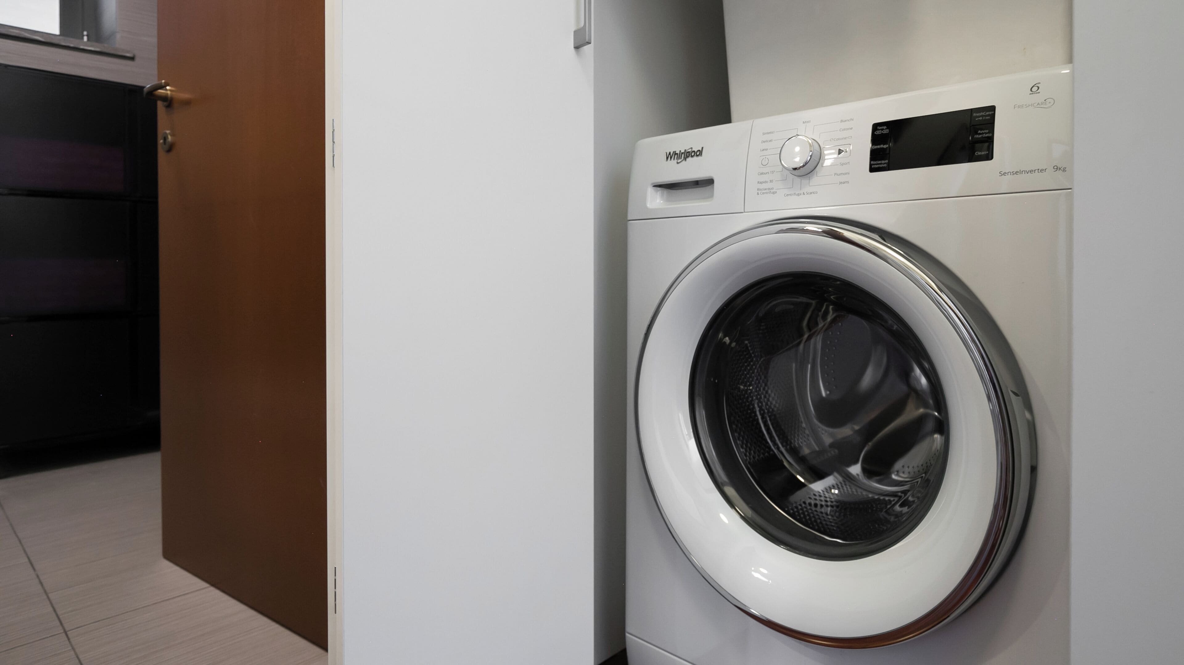 Uma máquina de lavar Whirlpool com painel de controle, num quarto com porta de madeira e soalho em azulejos.