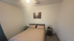 Uma cama de solteiro com cabeceira, duas mesas de cabeceira, uma luminária na parede e um quadro emoldurado sobre a cama.