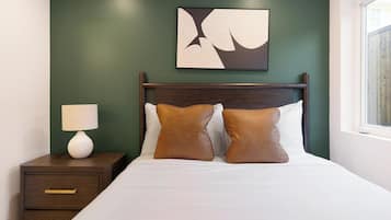 Uma cama bem arrumada com duas almofadas, uma mesa de cabeceira com abajur e uma obra de arte abstrata na parede.