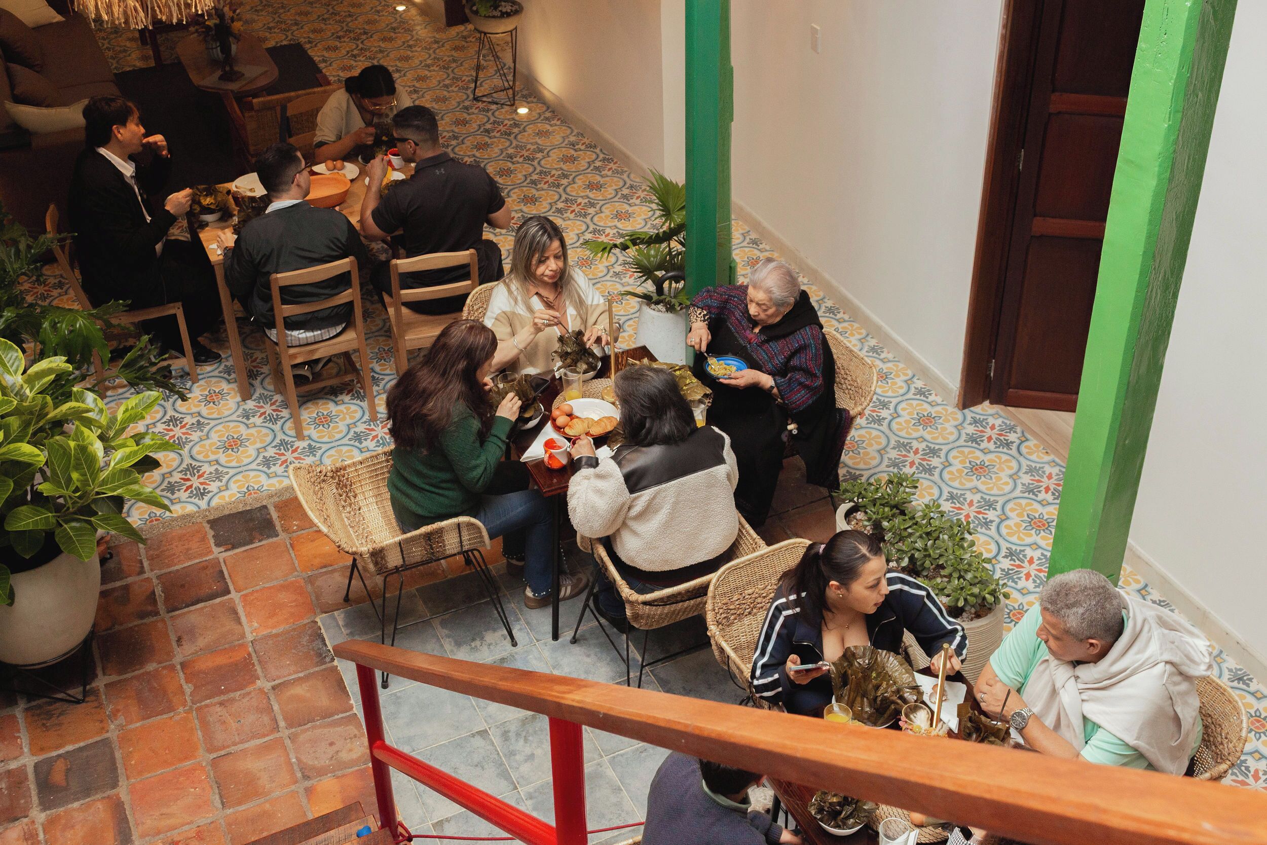 Uma sala de jantar com várias mesas e cadeiras, cercada por plantas em vasos e um piso com padrão.