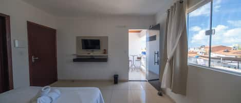 Quarto de hotel com cama, televisão, mesinha e varanda com vista para a cidade.