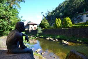 Uma estátua de um homem sentado à beira de um rio, com um muro de pedra e vegetação ao fundo.