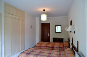 Quarto com duas camas, um guarda-roupa, uma mesinha, uma luminária e uma janela.